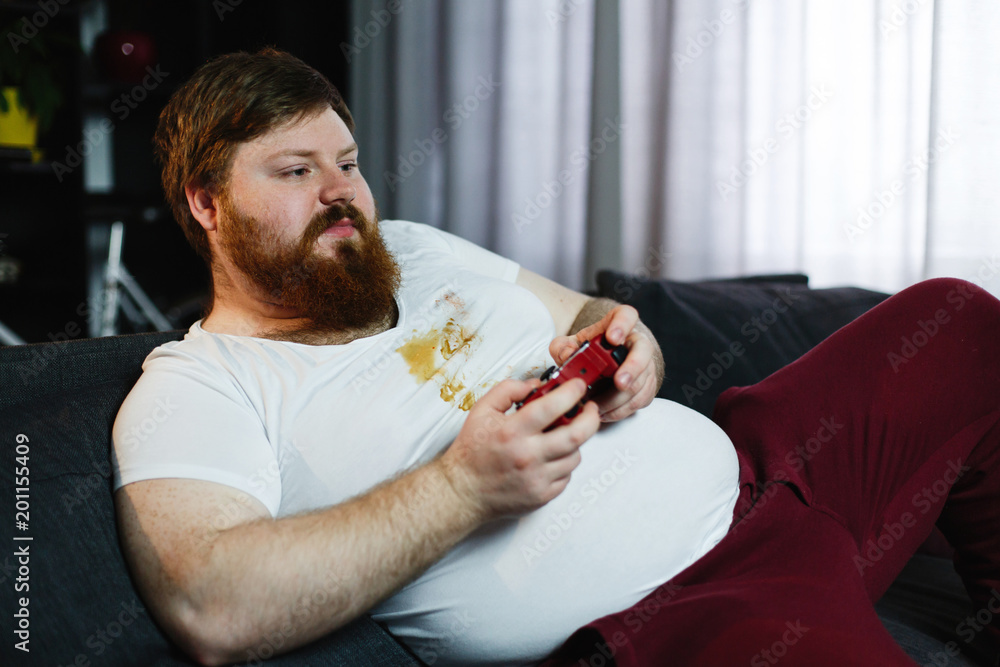 Happy fat man in dirty shirt plays video games drinking beer and eating ...