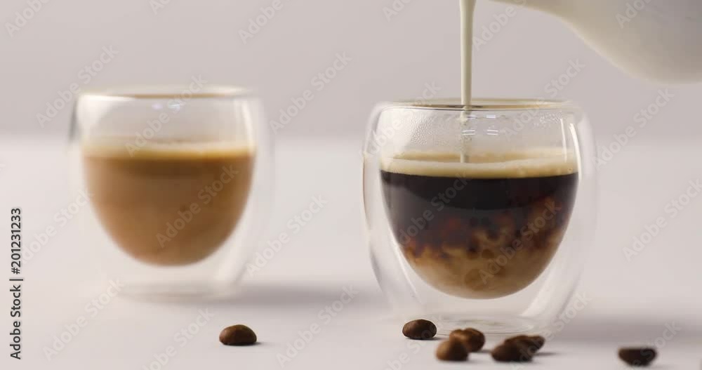 Pouring milk from jug in one of two cups with coffee on white ...