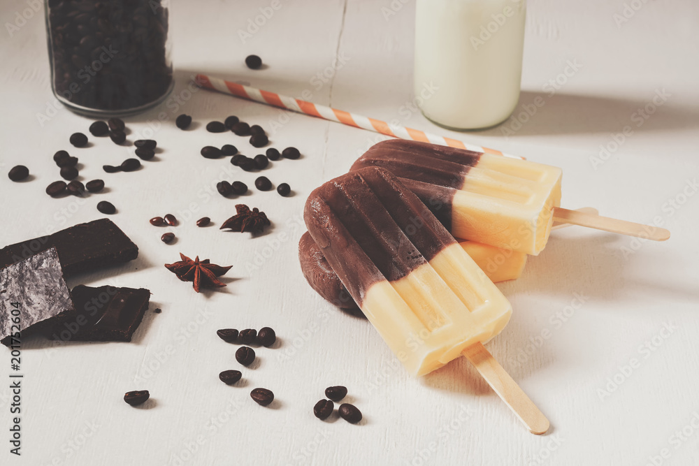 Two-colored ice cream on a stick, beige brown on a white table. Coffee ...