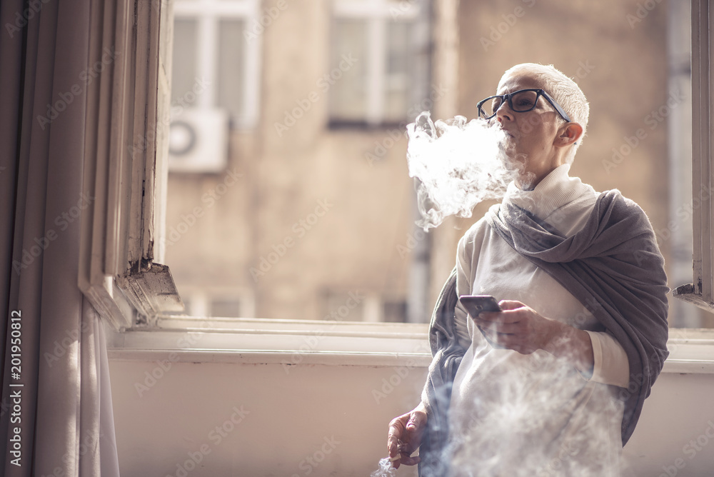 Senior elderly woman smoking a cigarette next to a big window in her ...