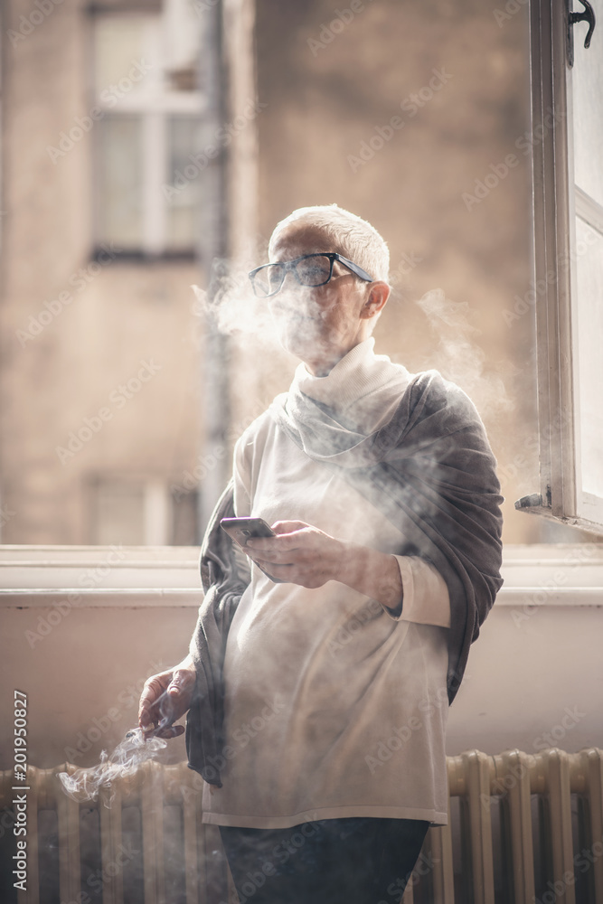 Senior elderly woman smoking a cigarette next to a big window in her ...