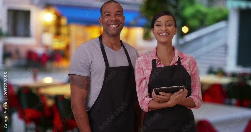 Welcoming warm African-American waiter and waitress invite camera over ...