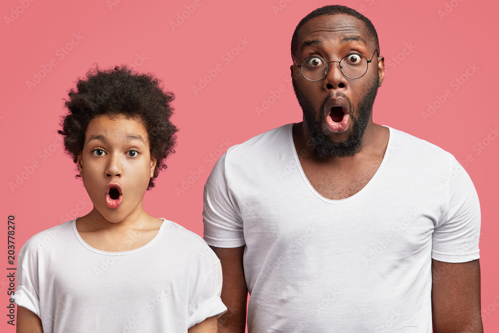 Indoor shot of shocked father and son stand next to each other, express ...