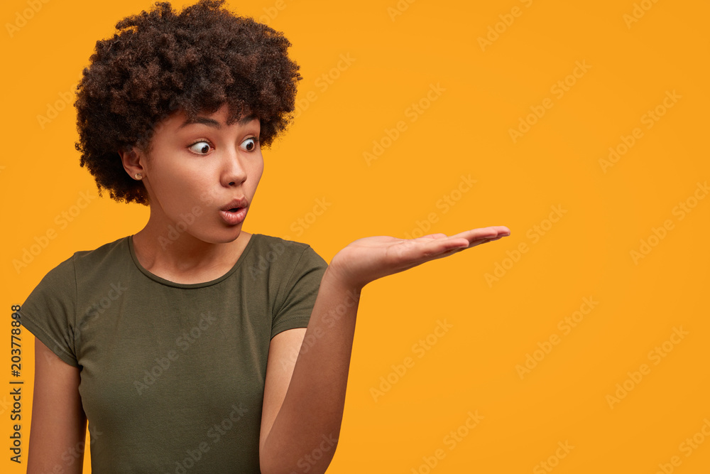 Horizontal shot of surprised African American woman indicates with palm ...