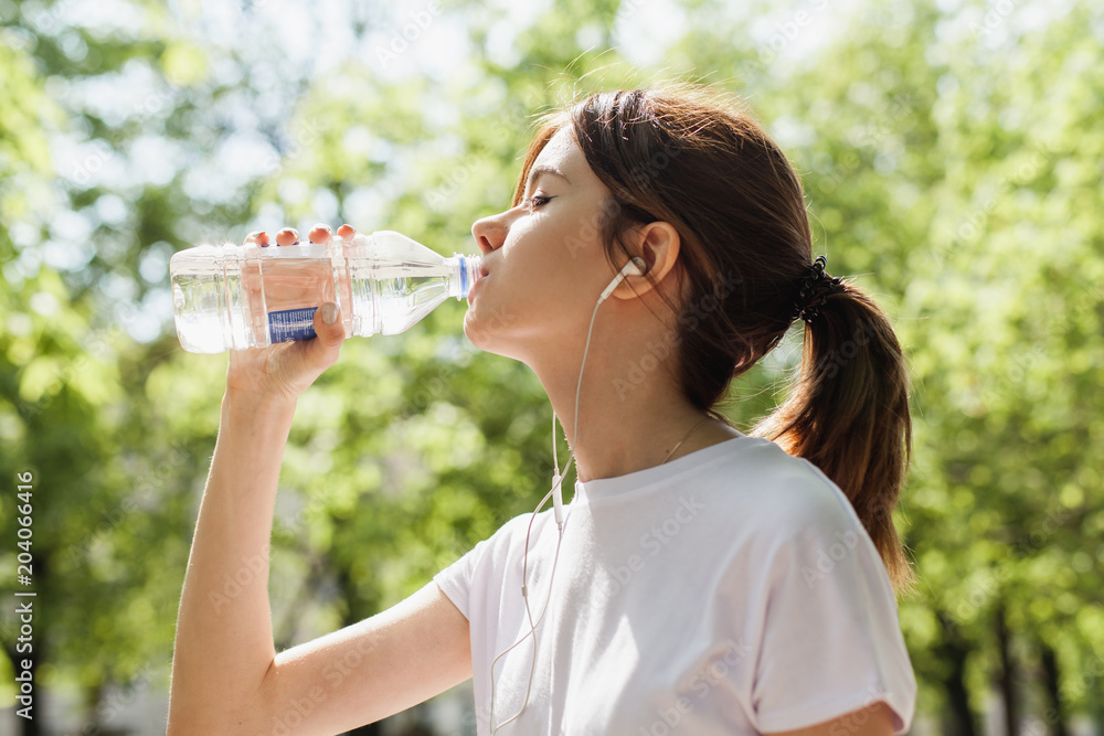 Young girl drinking water while walking in the park and listening to ...