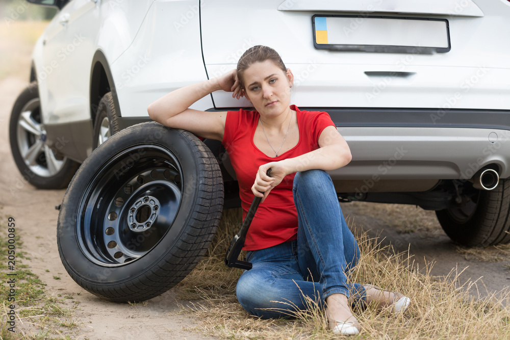 Young upset woman sitting on ground next to broken car on deserted road ...