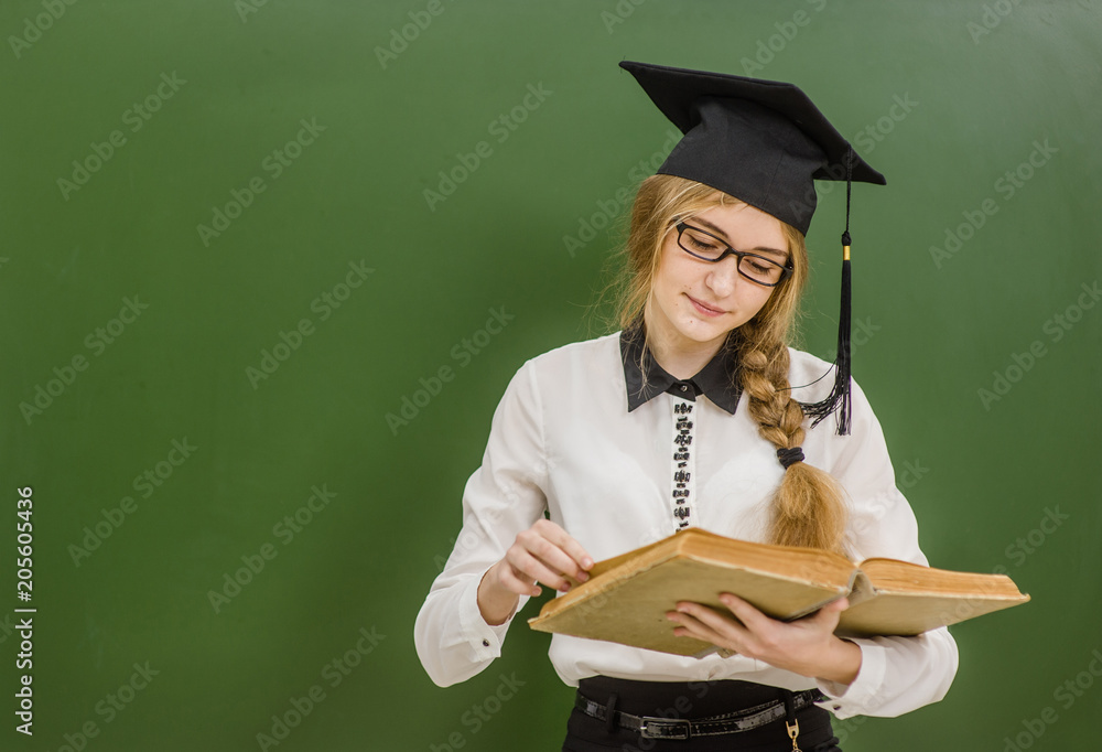 Teen girl in graduation cap with books standing near green chalkboard ...