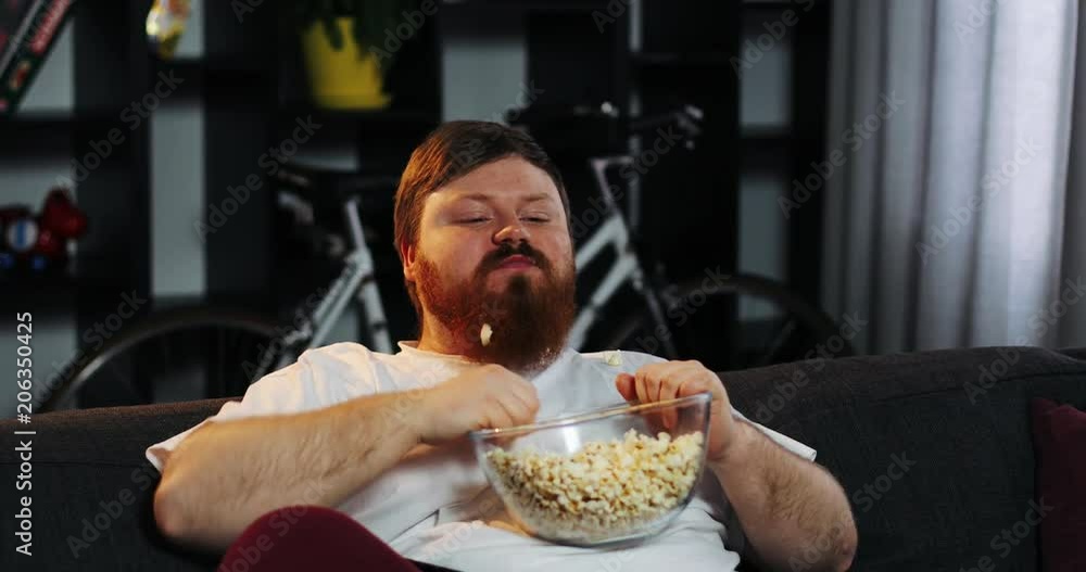 Smiling fat man with beard watches TV in the room and eats pop-corn at ...