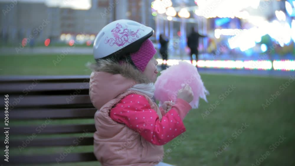 Beautiful little girl sitting on a bench in an amusement park, eating ...