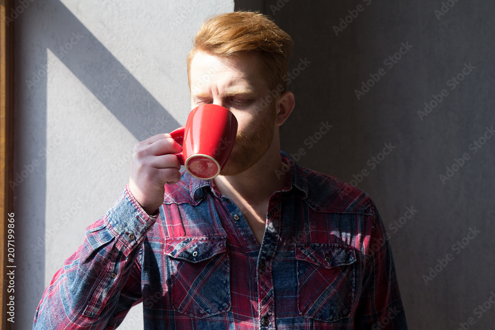 Handsome guy drinking from red cup covered in sunlight. Attractive man with ginger hair having ...