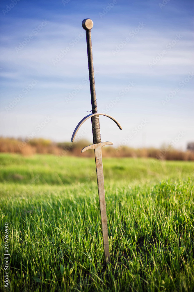 two-handed sword in the ground, against the background of a green field ...