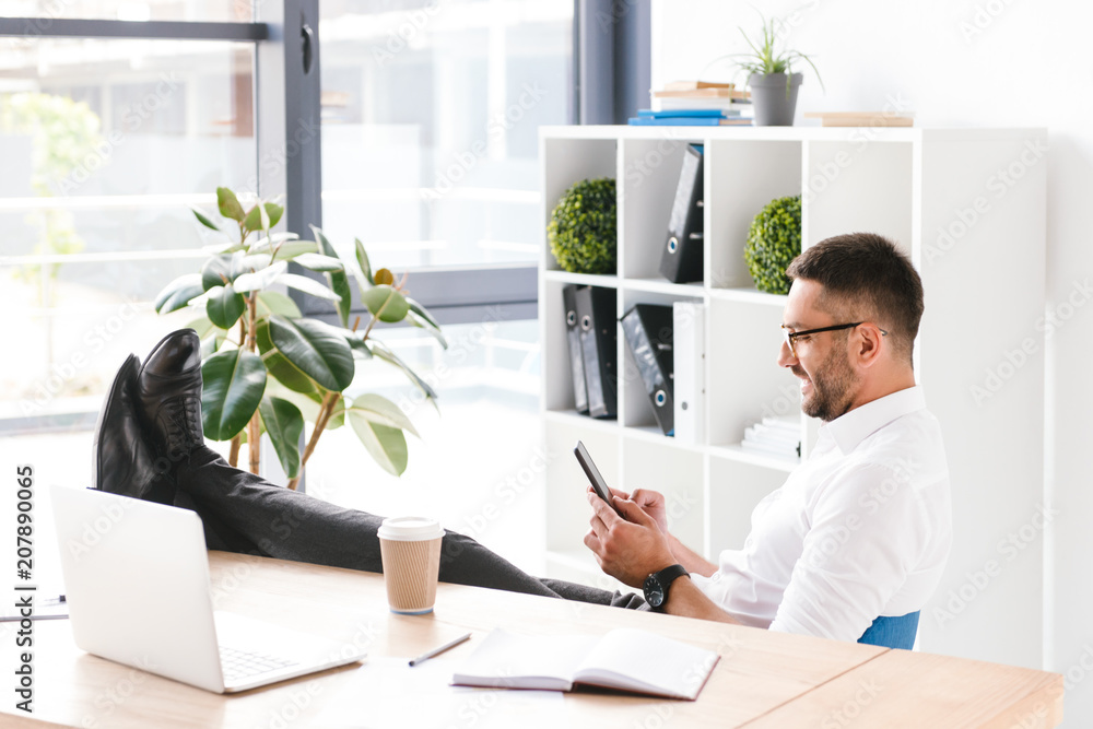 Image of happy handsome man in formal wear sitting in office with legs ...