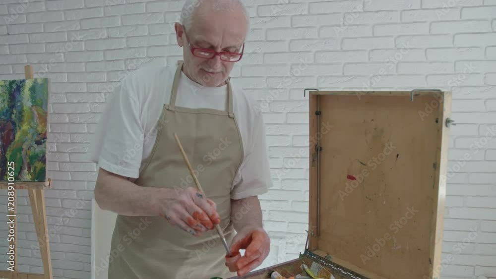 Old painter's hand taking paint with a brush, standing near big palette ...