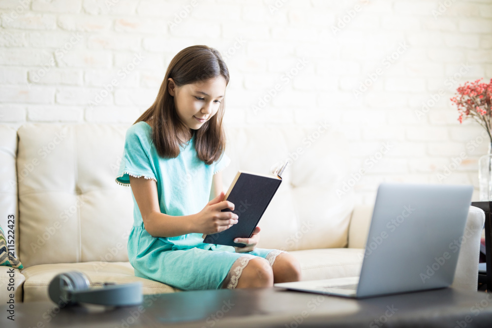 Young woman sitting on sofa reading book with laptop and earphone Stock ...