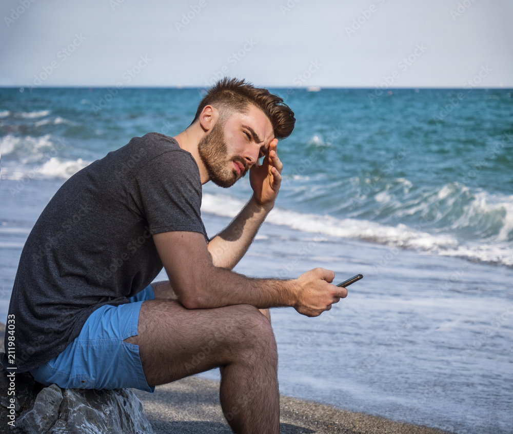 Handsome young man sitting on a beach, feeling lonely and sad or simply ...