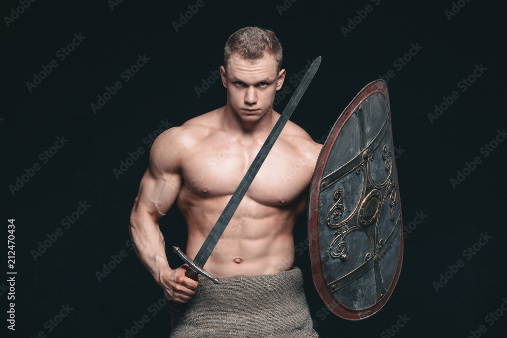 Bodybuilder man posing with a sword and shield isolated on black ...