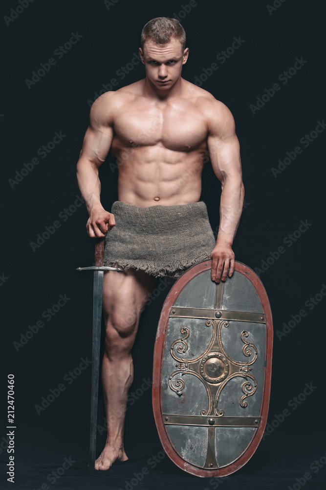 Bodybuilder man posing with a sword and shield isolated on black ...