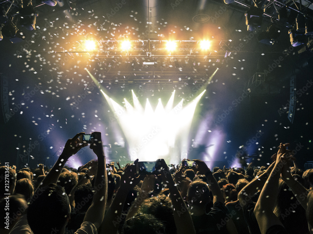 Rain of confetti falling on a concert crowd in front of a lit stage ...