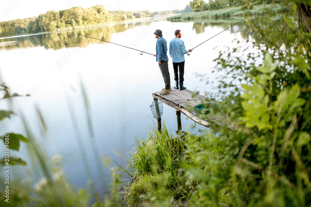Two male friends fishing together standing on the wooden pier during ...