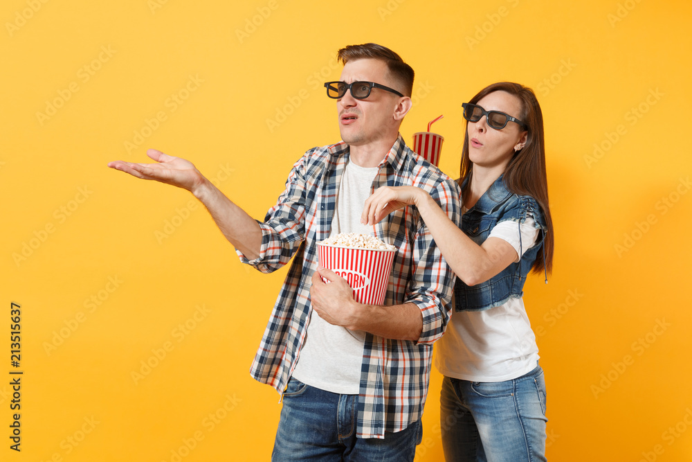 Young irritated couple woman man in 3d glasses watching movie film on date hold bucket of ...