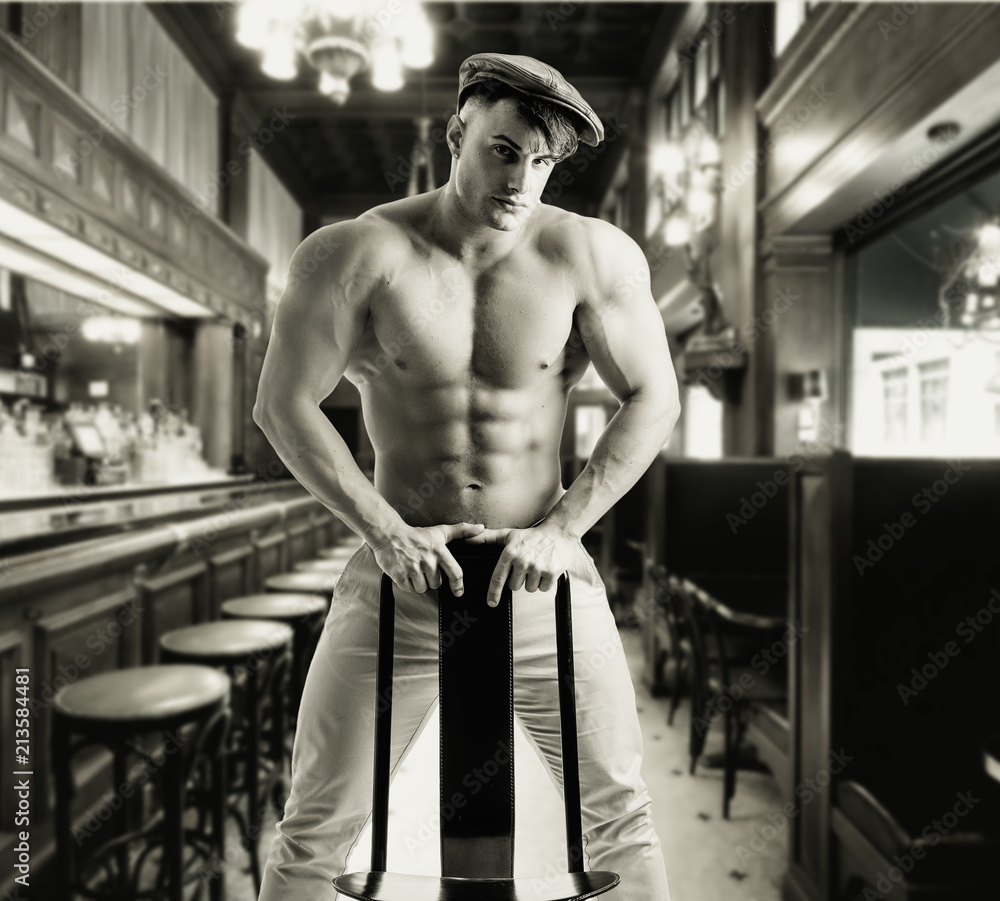 Black And White Shot Of Muscular Alluring Man Standing Shirtless With Chair In Bar Wearing Cap