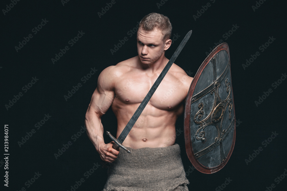 Bodybuilder man posing with a sword and shield isolated on black ...