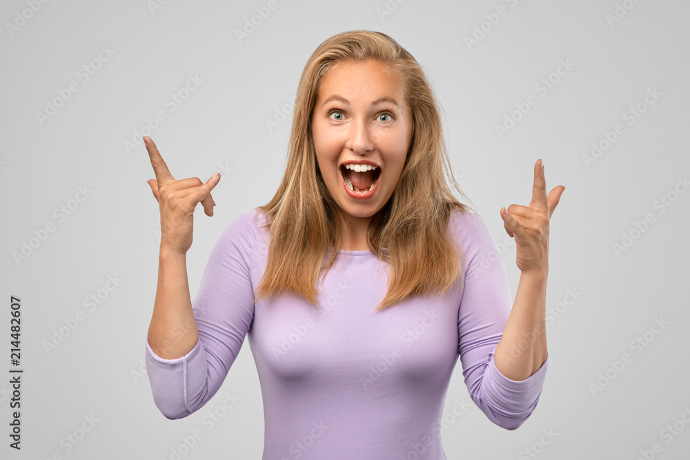 Crazy blond girl giving the Rock and Roll Sign gray studio background ...