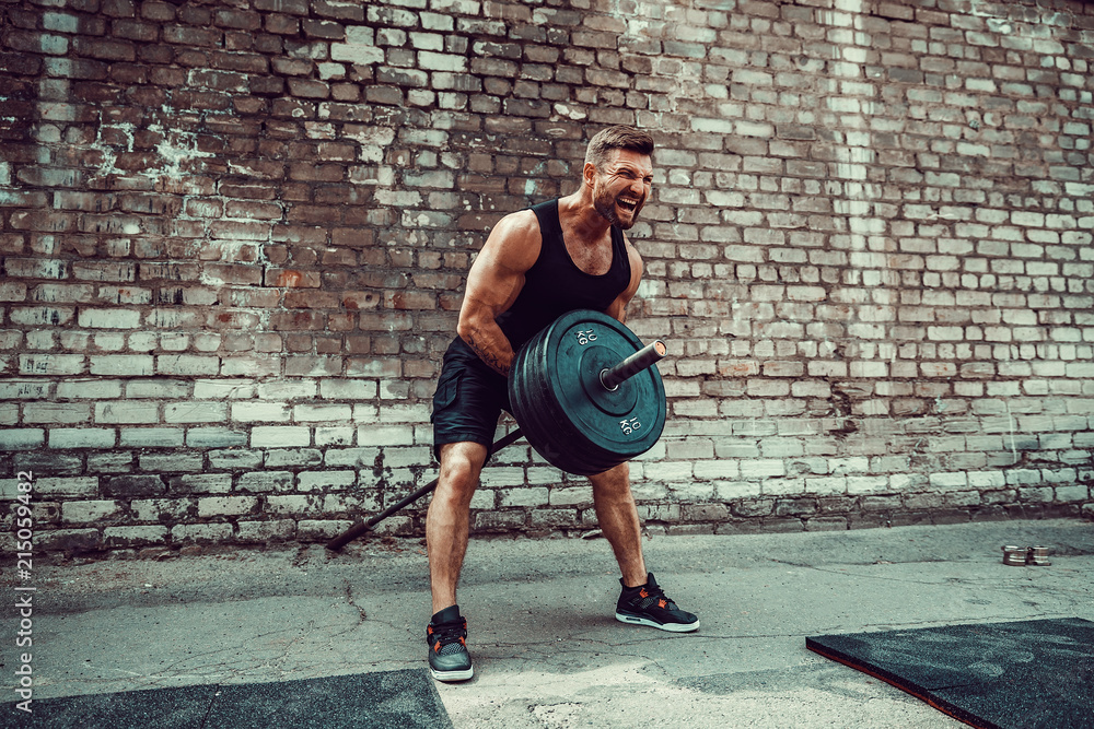 Athletic man working out with a barbell in front of brick wall ...