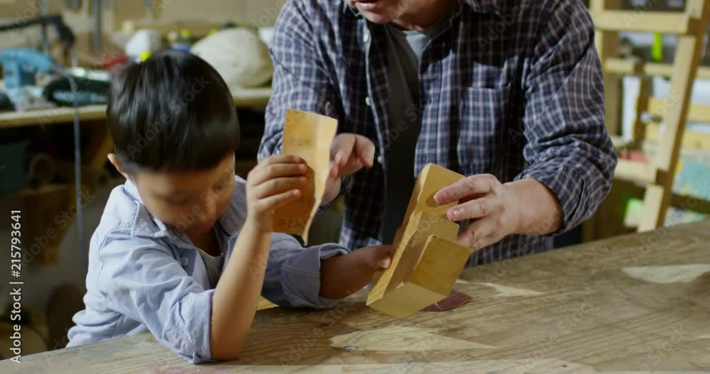 Handheld shot of cute Asian boy sanding wooden toy in carpentry ...