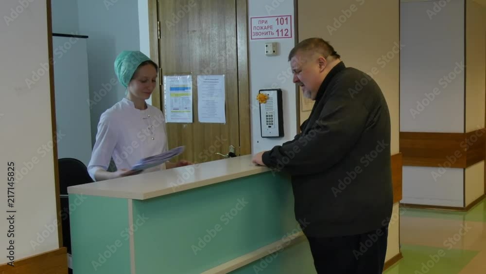 Nurse takes medical card from fat man in clinic. Girl in white lab coat ...