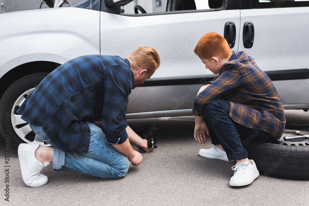father lifting car with floor jack for changing tire, son sitting on ...