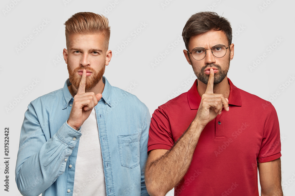 Two male friends with thick beards, keep fore fingers on lips, look ...