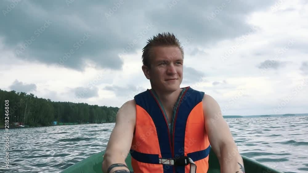 A young man in a boat rowing wooden oars on the water. Life jacket on ...