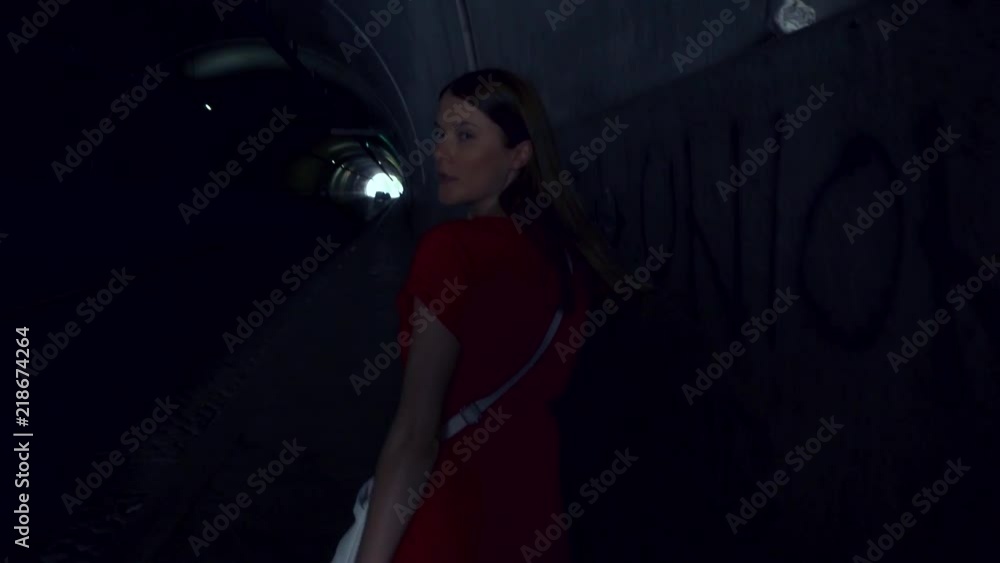 Back view of young woman in red dress walking alone in tunnel at night ...