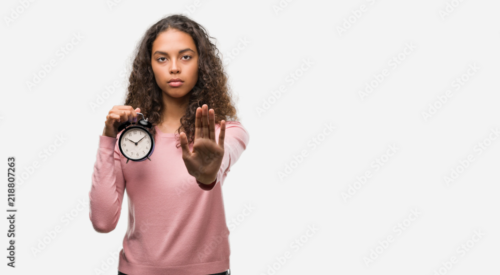 Young hispanic woman holding alarm clock with open hand doing stop sign ...