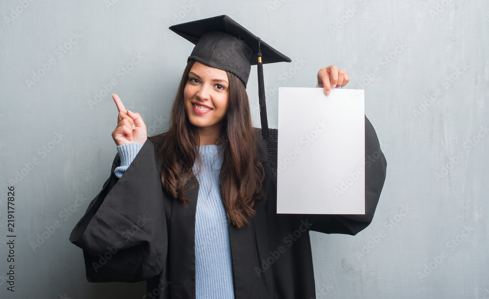 Young brunette woman over grunge grey wall wearing graduate uniform ...