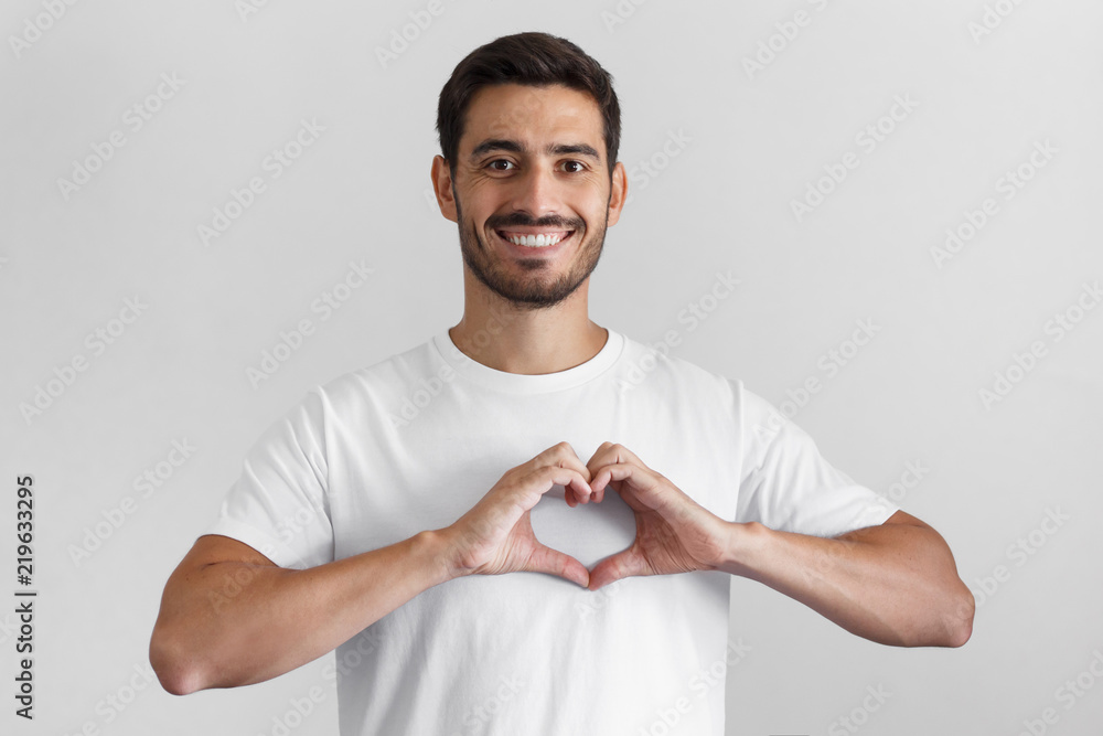 Portrait of smiling young man keeps hands on chest in heart shape sign ...