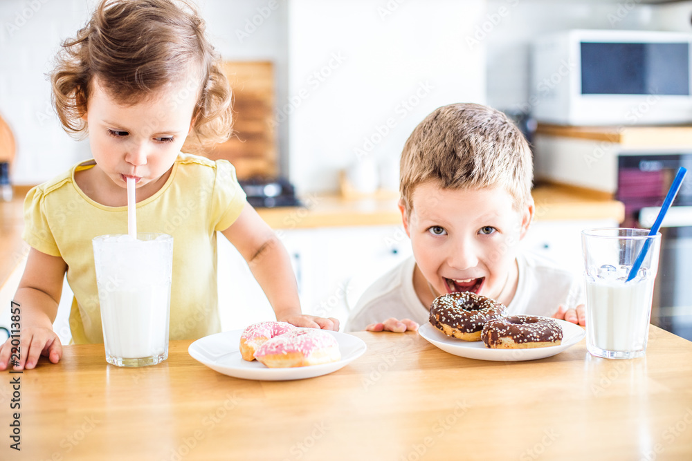 Children eating donuts and drinking milk on the white kitchen at home ...