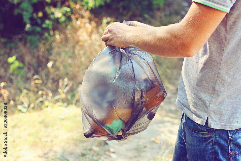 hand holds against a forest full of garbage a large black plastic bag ...