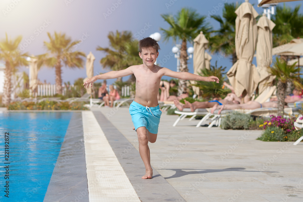 Portrait of happy Caucasian boy running along swimming pool at resort ...