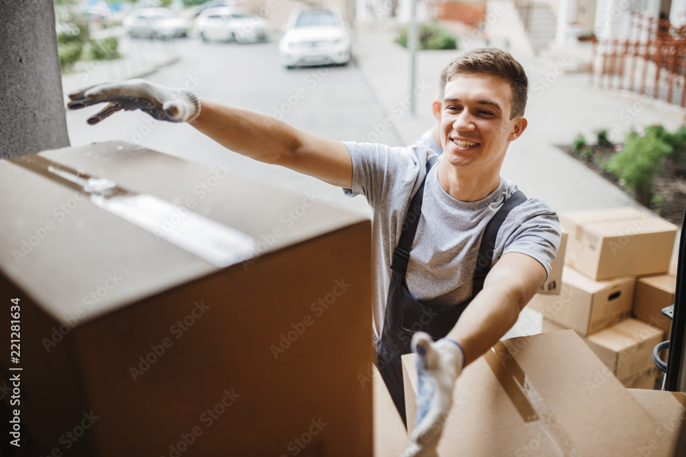 A young handsome smiling mover wearing uniform is reaching for the box ...