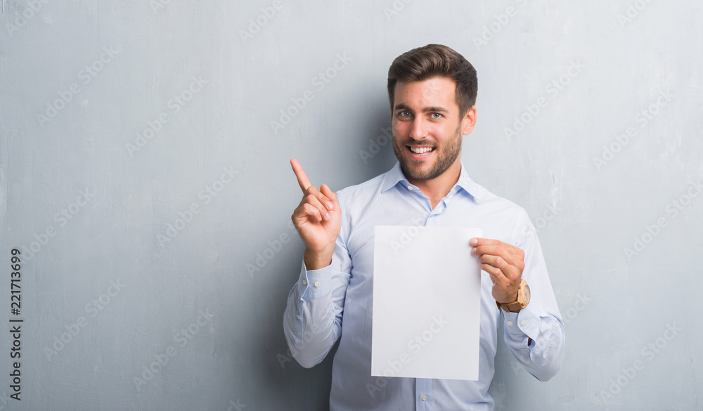 Handsome young man over grey grunge wall holding blank paper sheet ...