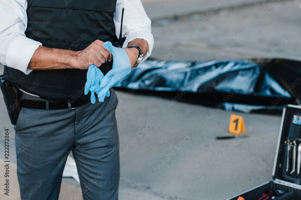 cropped image of policeman with gun in holster putting on latex gloves ...