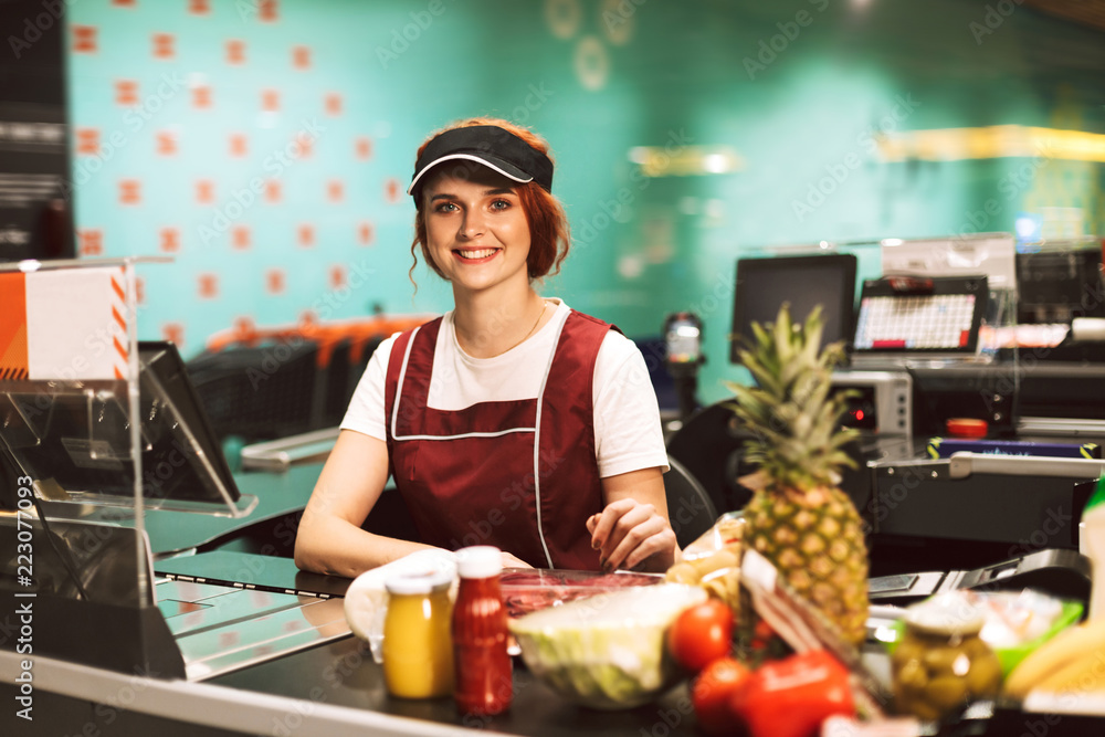Young beautiful cashier in uniform happily looking in camera while ...
