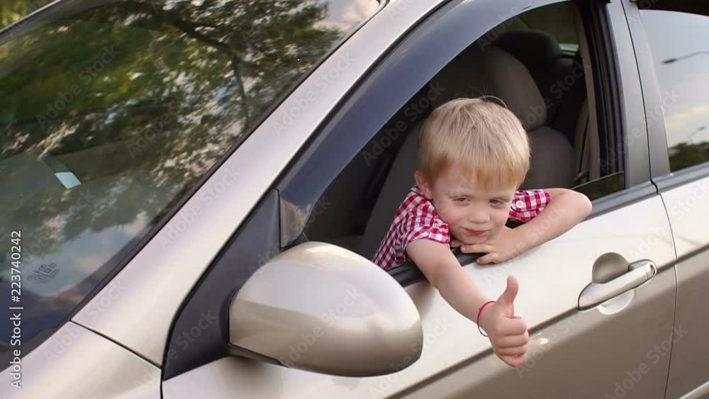 A little happy boy is sitting behind the wheel of a car and shows his ...