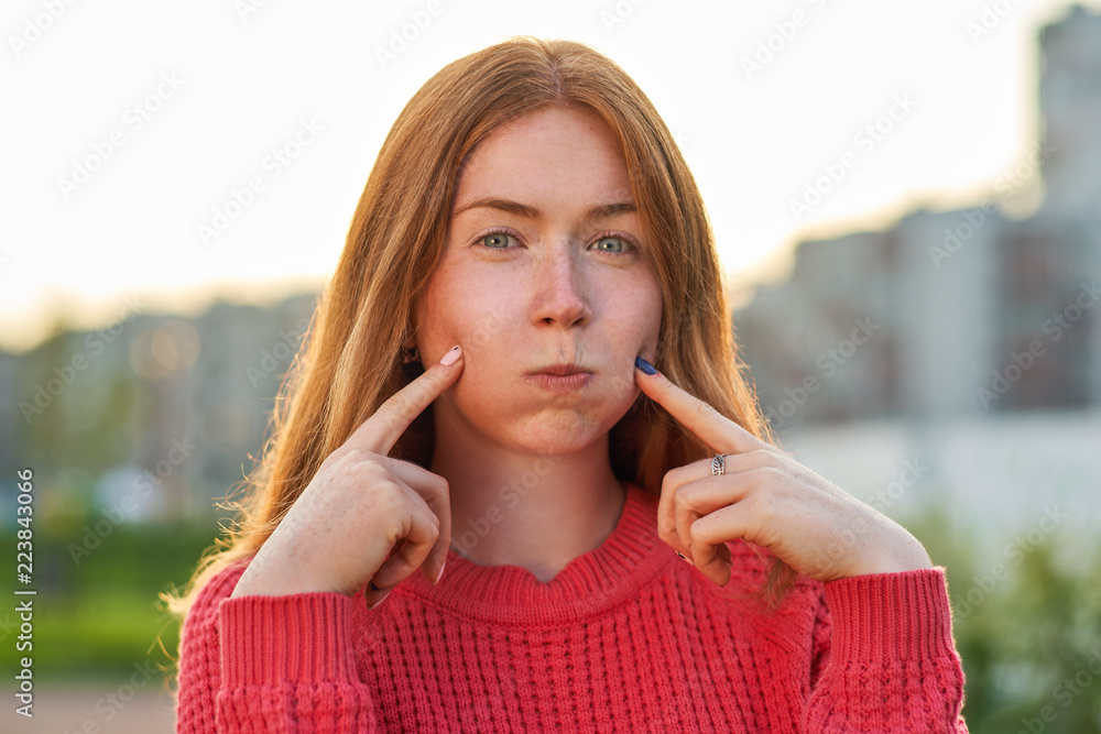 A funny red-haired girl inflates her cheeks and pokes her index fingers ...