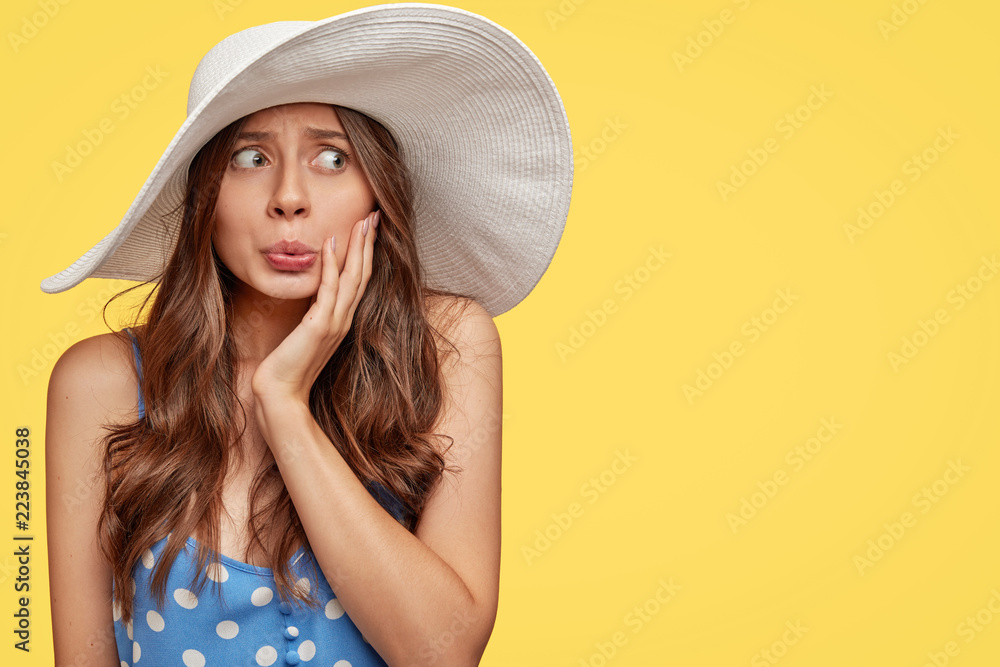 Puzzled nervous young lady in fashionable summer hat, presses lips and ...