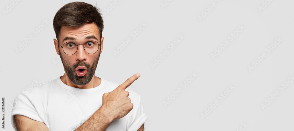 Photo of shocked European man with thick bristle points at blank copy ...
