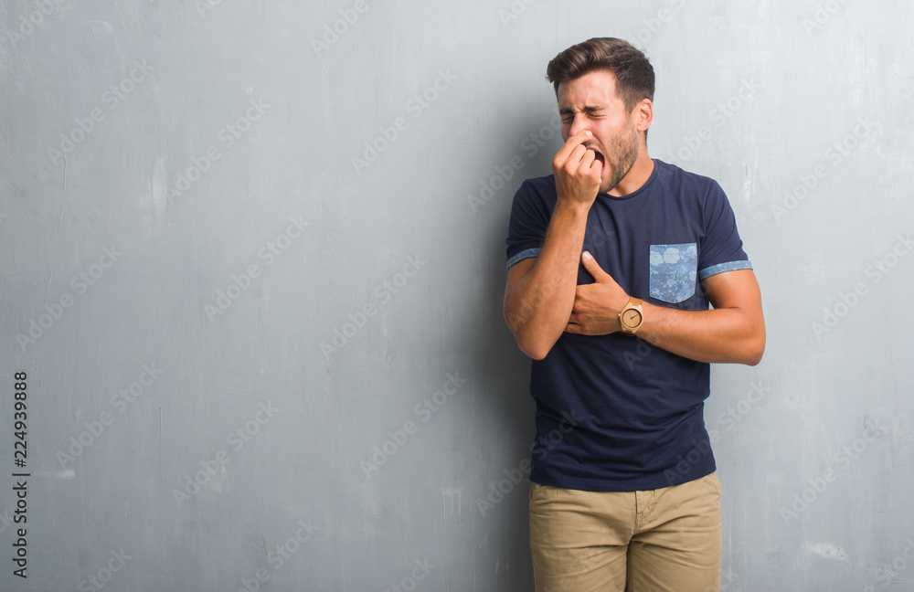 Handsome young man over grey grunge wall smelling something stinky and ...