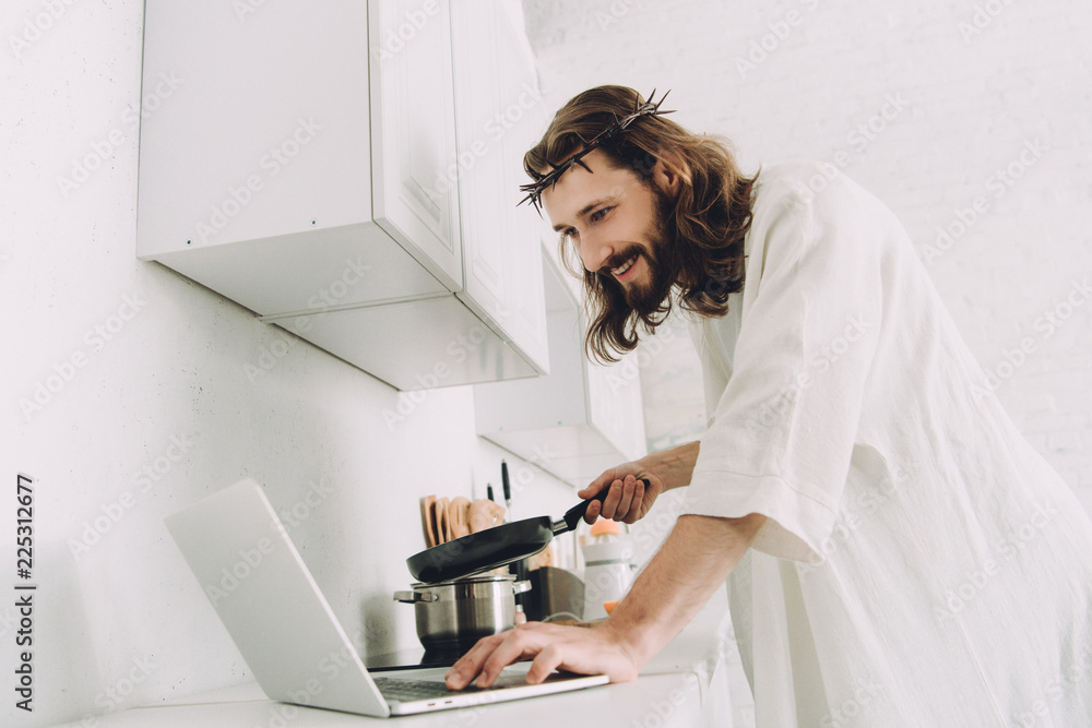 smiling Jesus using laptop while cooking with frying pan in kitchen at ...